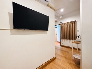 Quarto com televisão montada na parede, piso de madeira, janela com cortinas e uma pequena mesa com chaleira.