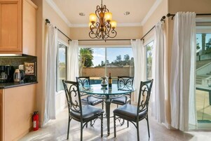 Uma sala de jantar com mesa de vidro, quatro cadeiras, um candelabro e vista para uma área residencial através de grandes janelas.