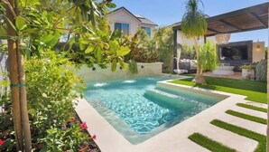 Uma piscina com um recurso de água azul cristalina, rodeada por vegetação bem cuidada e uma área de estar moderna ao ar livre.