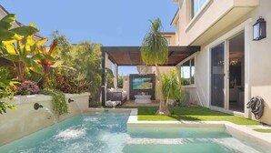 Piscina com pergolado e área de estar externa, rodeada por vegetação exuberante e um gramado bem cuidado.