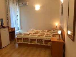 Quarto com cama, abajures, escrivaninha, cadeira, mini-geladeira e janela com cortinas.