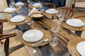 Uma mesa de jantar posta com pratos brancos, talheres e copos de vidro transparente, rodeada por jogos americanos trançados e cadeiras de madeira.