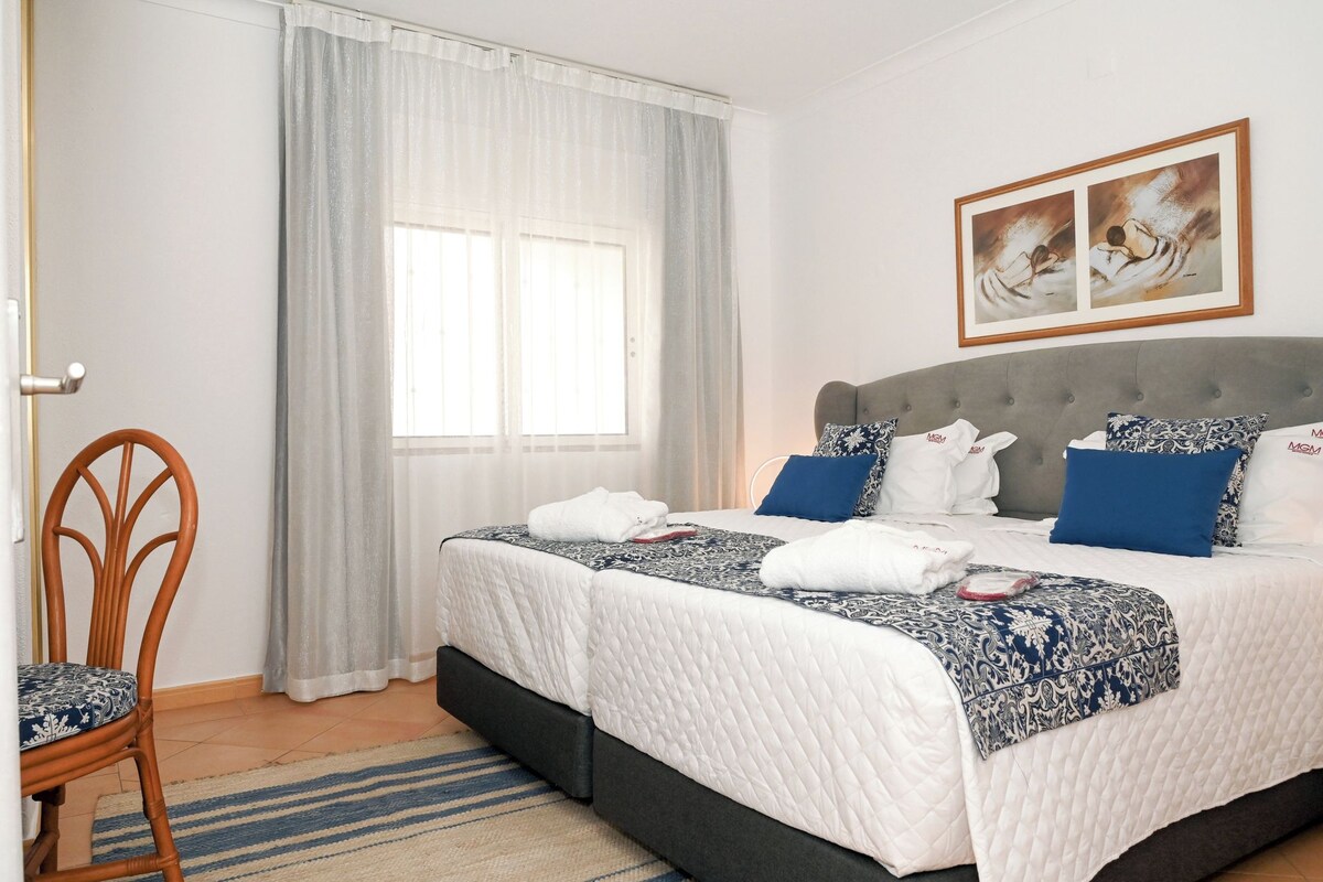 Quarto de hotel com cama, uma cadeira, janela com cortinas e um quadro na parede.