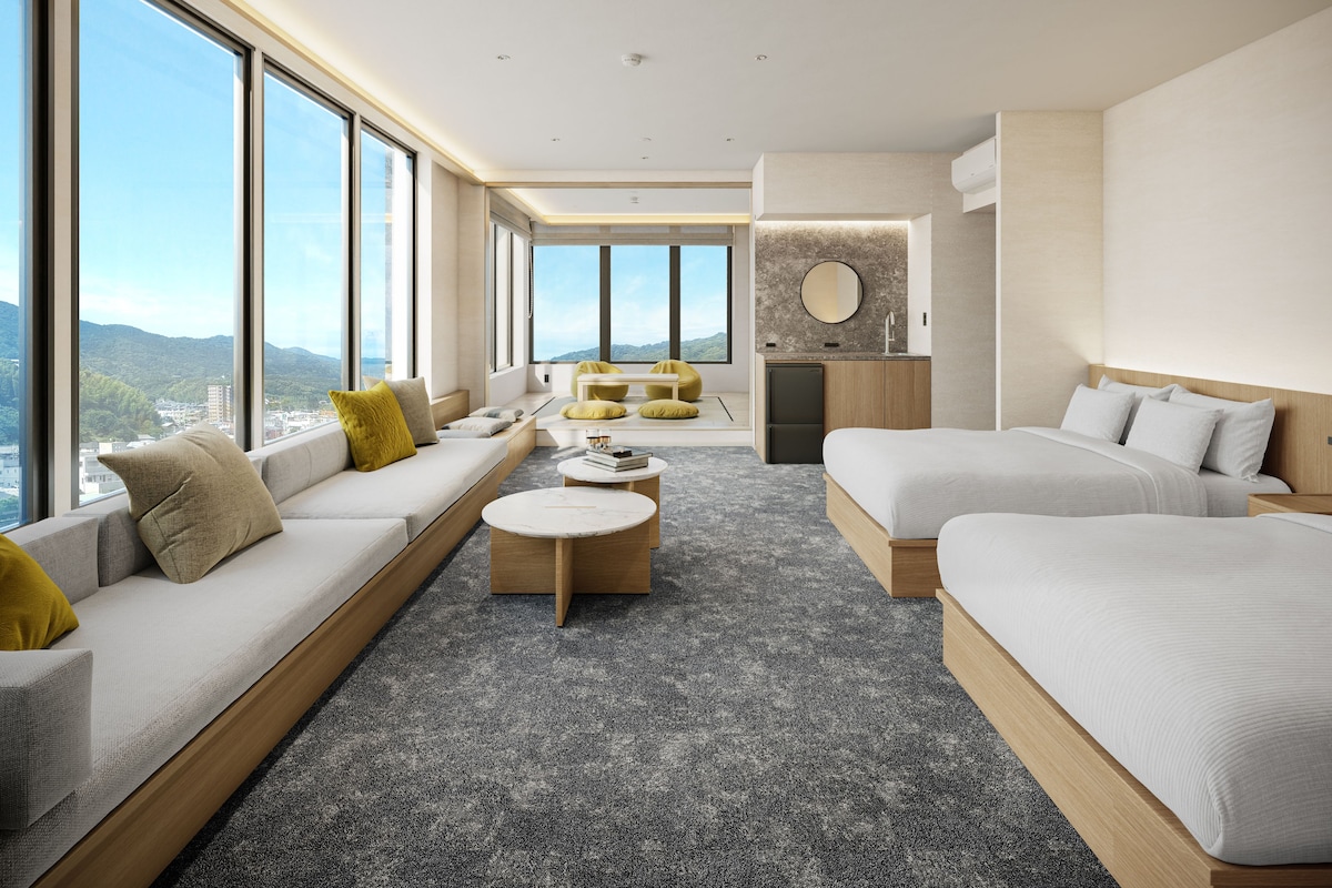 Quarto de hotel moderno com uma grande janela, um sofá, uma cama, uma pequena cozinha e vista para a cidade.