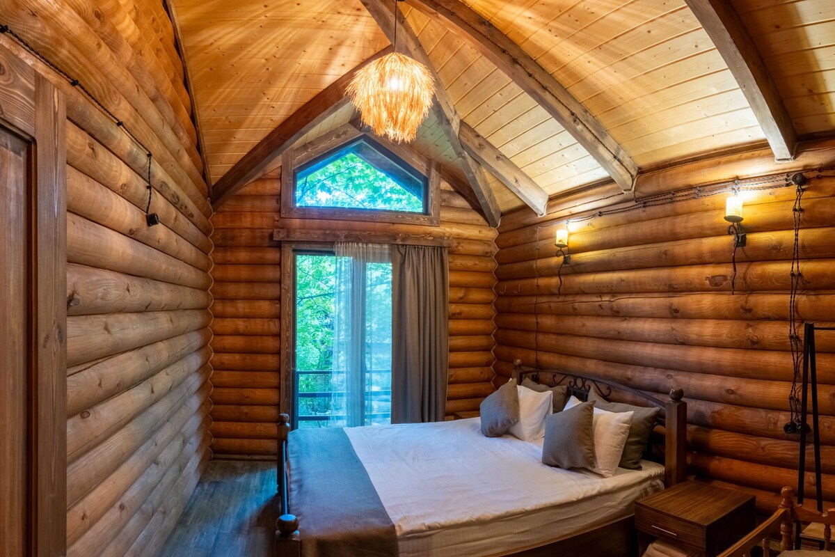 Quarto de cabana de madeira com cama, janela com cortinas, luminária pendente e mesa de cabeceira de madeira.