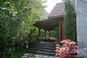 Um gazebo de madeira com parede de pedra, rodeado por vegetação exuberante e um caminho de pedra.
