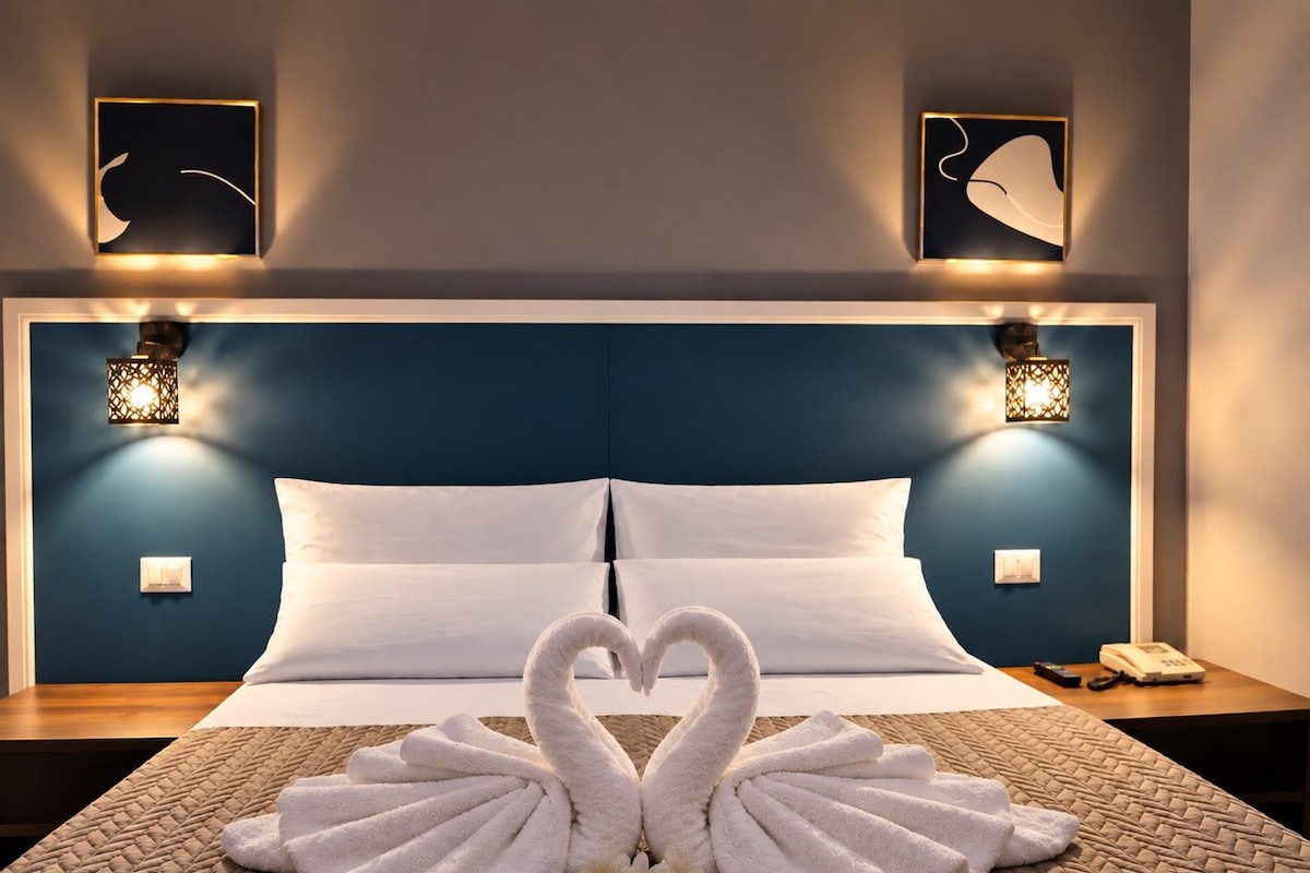 Quarto de hotel com uma cama grande, duas luminárias fixadas na parede e arranjo decorativo de toalhas em formato de cisne.