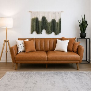 Um sofá de couro marrom com quatro almofadas, uma luminária de chão, uma planta presa na parede e um tapete texturizado.