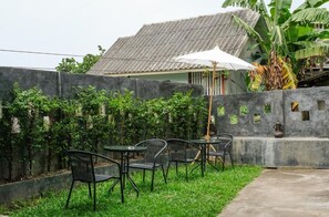 Uma área externa com mesas e cadeiras, um muro de concreto e um edifício com telhado de metal corrugado ao fundo.