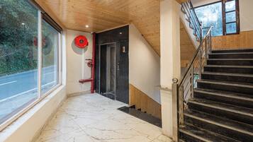 Um corredor com piso de mármore, um alarme de incêndio vermelho e uma escada com degraus de madeira.
