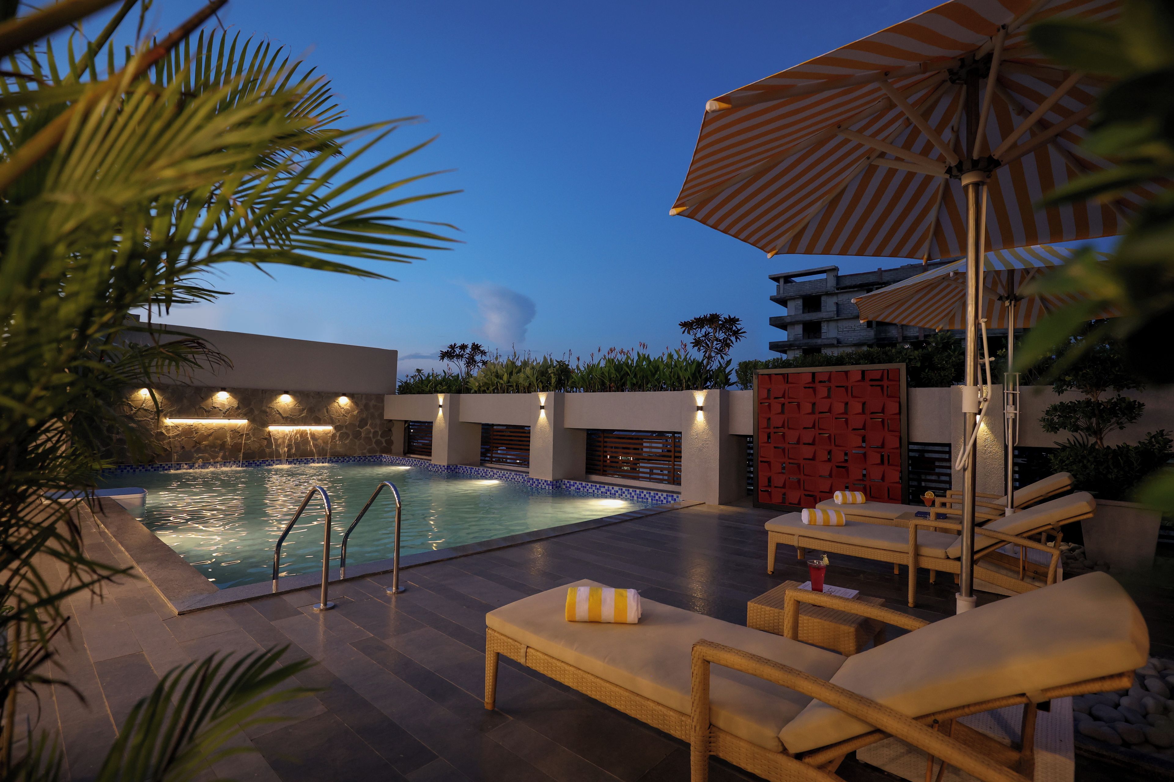 Área de piscina com espreguiçadeiras e um guarda-sol, cercada por plantas e uma parede de pedra.