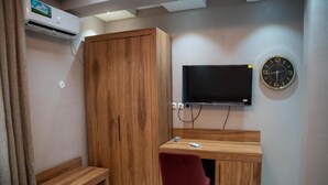 Um quarto com uma cama, guarda-roupa de madeira, escrivaninha com cadeira, televisão e relógio de parede.