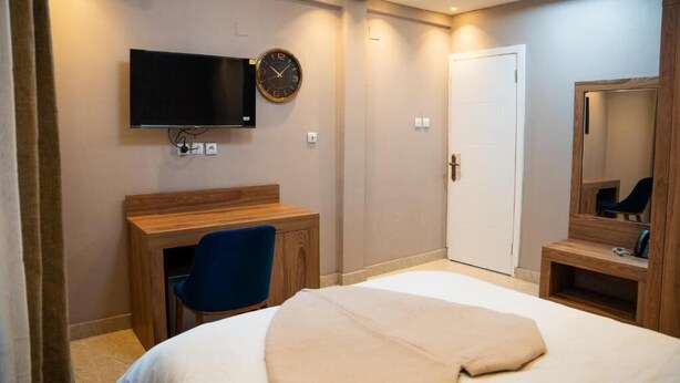 Quarto de hotel com cama, escrivaninha, cadeira, televisão, relógio e porta.