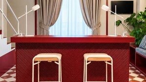 Um quarto com piso xadrez, um balcão vermelho com dois banquinhos, uma televisão e uma janela com cortinas.