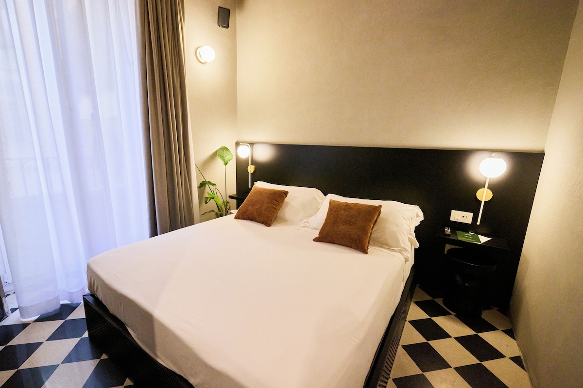 Quarto de hotel com uma cama grande, duas almofadas, uma mesa de cabeceira com abajur e chão xadrez.