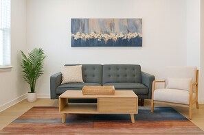 Uma sala de estar moderna com um sofá cinza, uma mesa de centro de madeira, uma poltrona branca e um quadro abstrato grande na parede.