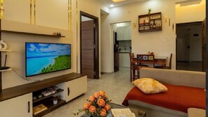 Uma sala de estar moderna com uma televisão de tela plana, um sofá, uma mesa de centro com um vaso de flores e um espaço de jantar com mesa e cadeiras.
