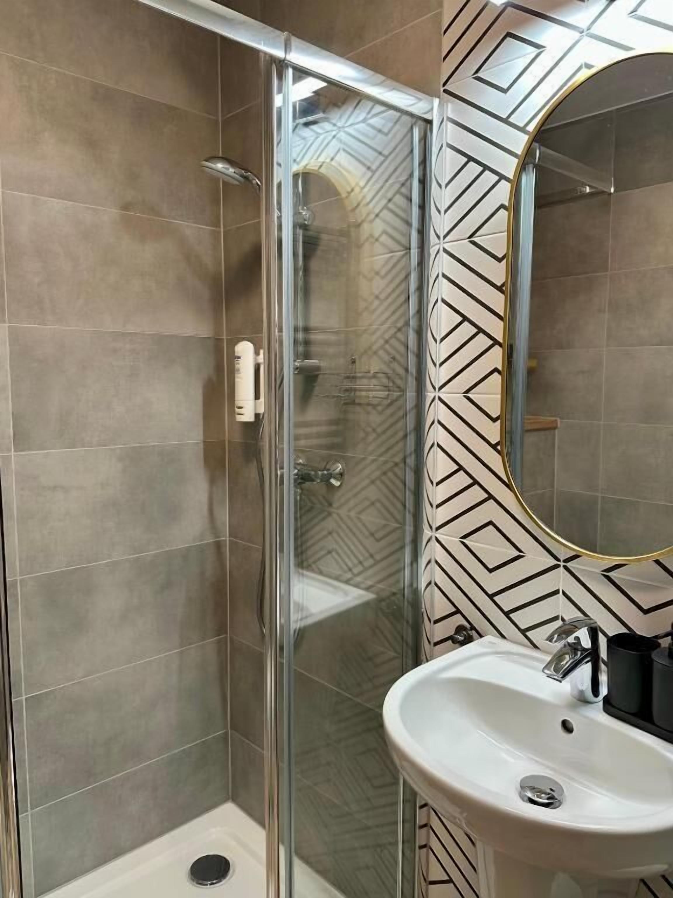 Um chuveiro com porta de vidro, uma pia com espelho e azulejos estampados nas paredes.