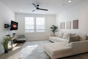 Uma sala de estar moderna com um sofá bege, uma TV fixada na parede, um ventilador de teto e uma janela grande com persianas.