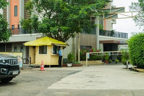 Uma cabine telefônica amarela com uma pessoa em frente, um carro preto estacionado por perto e um edifício com uma placa que diz 'MANAS'.