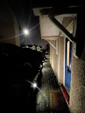 Vista noturna de um caminho estreito que leva a um edifício com porta azul, iluminado por postes de luz.