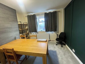 Um quarto com uma mesa de madeira, duas cadeiras, uma cadeira de escritório preta, um radiador e uma janela com cortinas.
