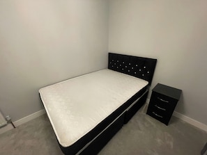Uma cama de solteiro com cabeceira, mesa de cabeceira e um móvel com gavetas num quarto com paredes lisas e chão coberto de carpete.