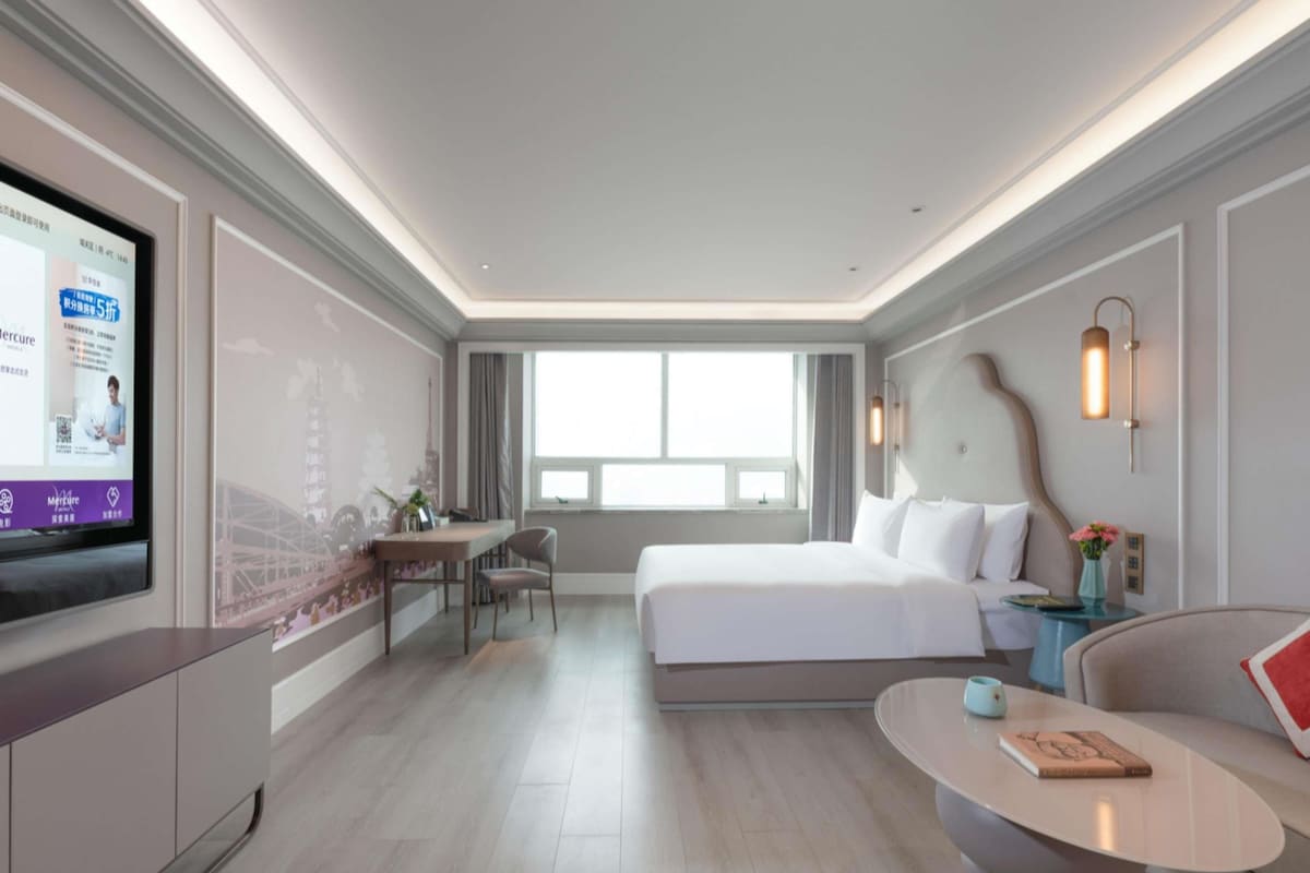 Quarto de hotel com cama, escrivaninha, cadeira, televisão e janela.