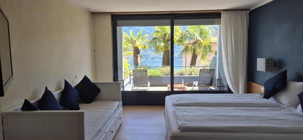Quarto de hotel com varanda, uma cama, área de estar e vista para palmeiras e um corpo d'água.