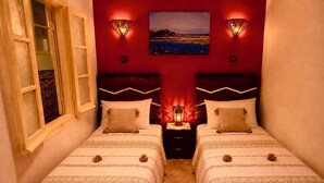 Quarto de hotel com duas camas, mesa de cabeceira, luminária na parede e pintura de uma paisagem costeira.