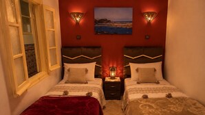 Quarto de hotel com duas camas, uma mesinha de cabeceira, um abajur e uma pintura de uma paisagem costeira.