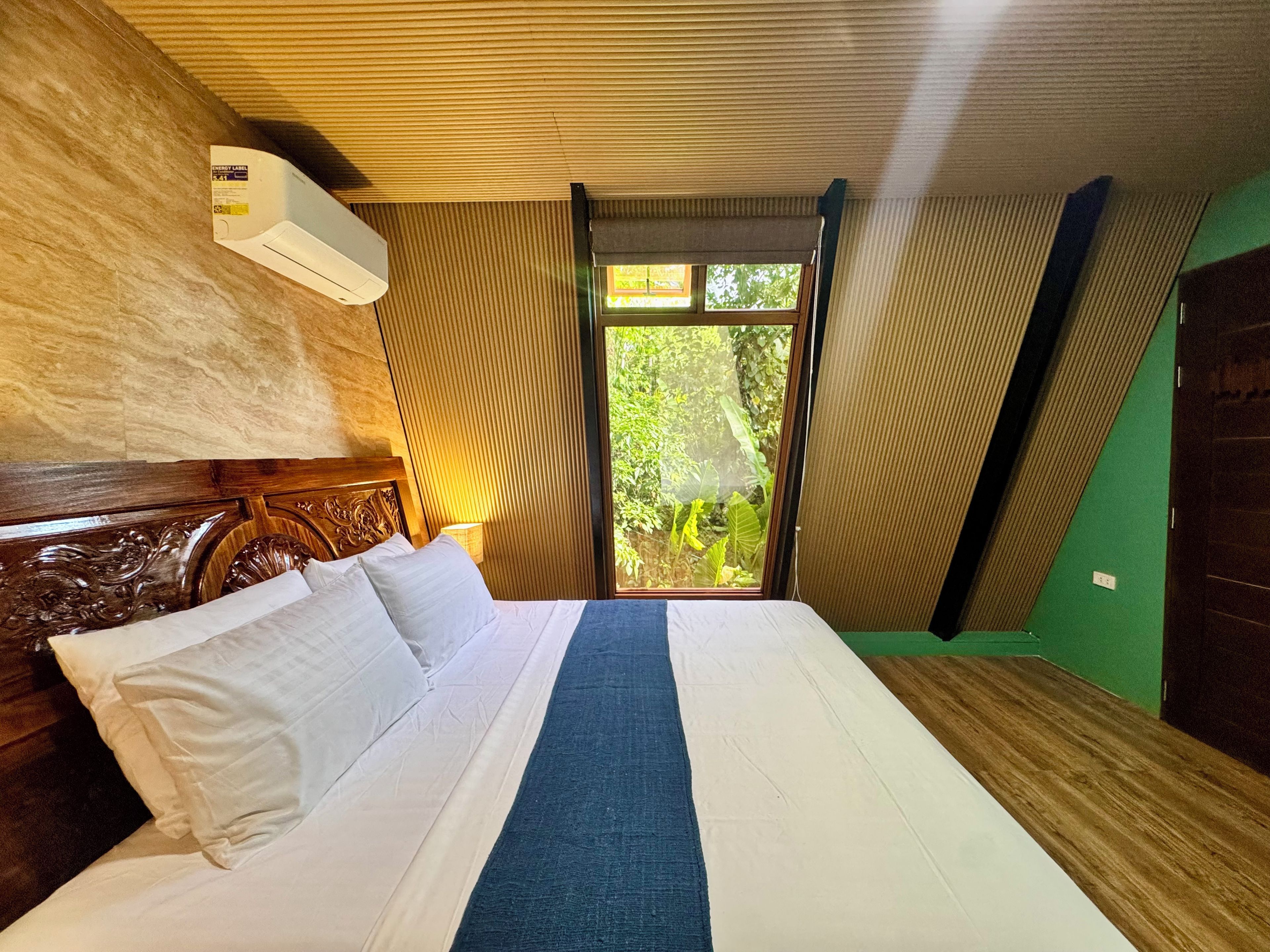 Quarto de hotel com uma cama, janela com vista para vegetação e teto de madeira.