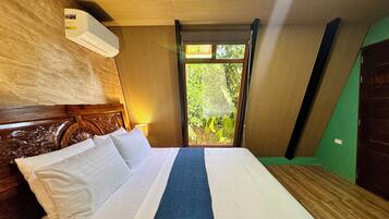 Quarto de hotel com uma cama, janela com vista para vegetação e teto de madeira.