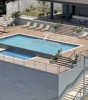 Uma piscina retangular com borda azulejada, rodeada por um espaço de deck com espreguiçadeiras e uma escadaria que leva a um nível superior.