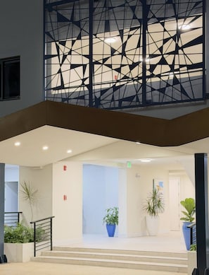 Entrada de um edifício moderno com fachada de vidro geométrica, uma escadaria que leva ao andar superior e plantas em vasos.
