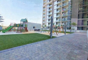 Um parquinho com um escorregador e estruturas de escalada, cercado por uma área pavimentada e um prédio residencial com varandas.