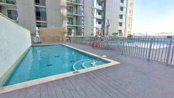 Uma piscina com um deck revestido de azulejos, cercada por uma grade de metal, e adjacente a um edifício alto com varandas.