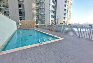 Uma piscina com um deck revestido de azulejos, cercada por uma grade de metal, e adjacente a um edifício alto com varandas.