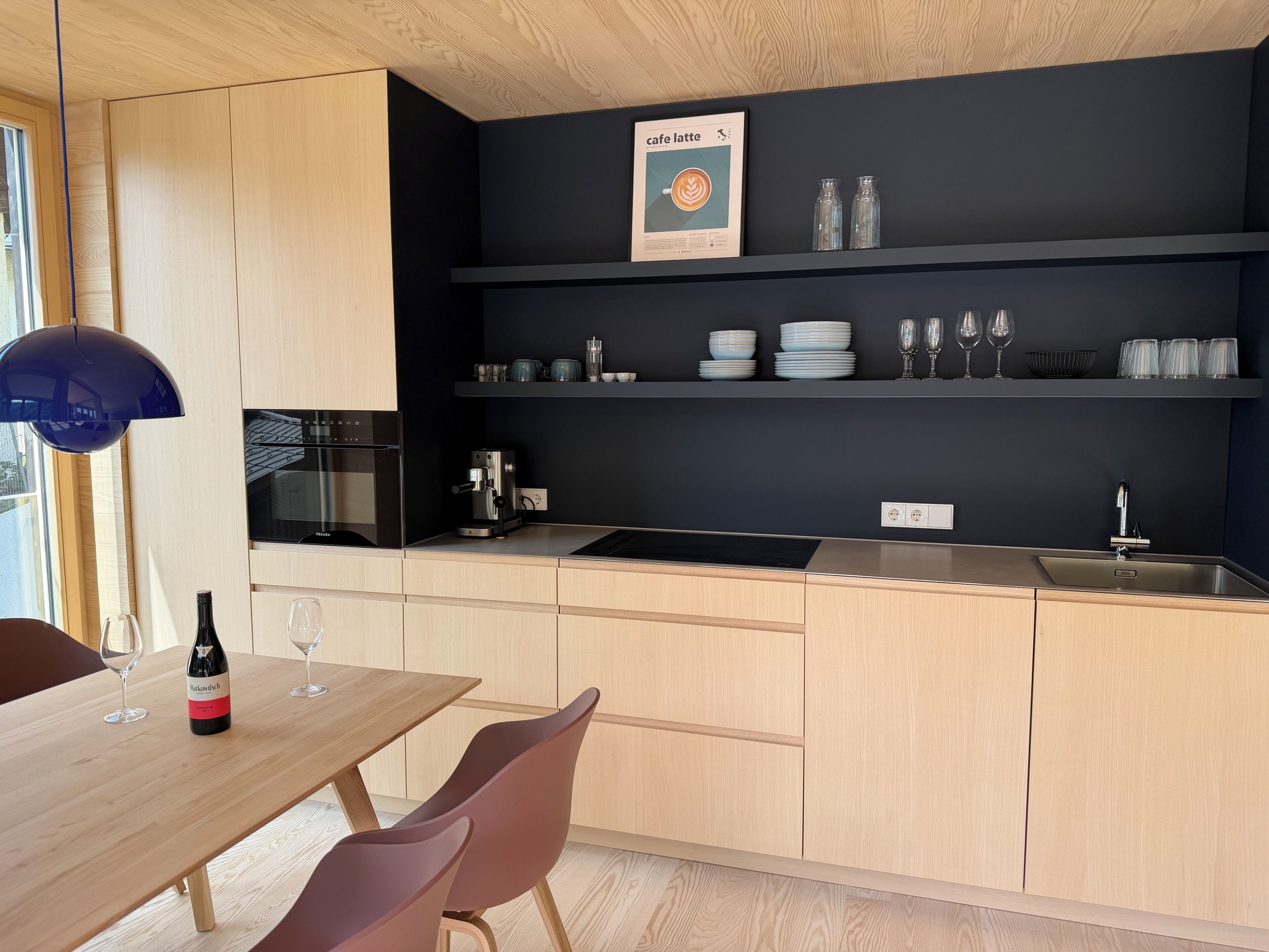 Cozinha moderna com armários de madeira, painel de fundo preto, cafeteira e área de jantar com mesa e cadeiras.