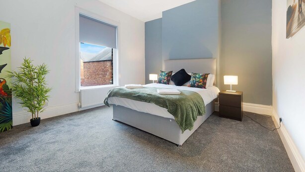 Um quarto com cama, janela, radiador, mesa de cabeceira, abajur, planta e um quadro na parede.