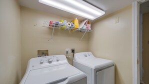 Uma lavanderia com duas máquinas de lavar de abertura frontal, uma prateleira com produtos de limpeza e uma luminária no teto.