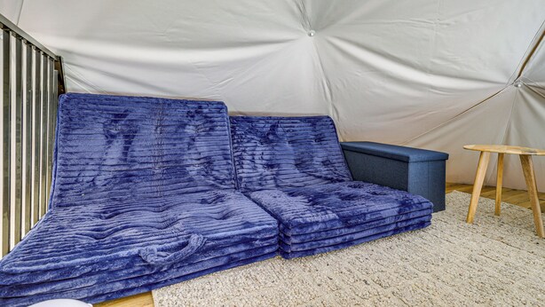 Um quarto com um sofá azul, um banquinho de madeira e uma estrutura em forma de tenda branca ao fundo.