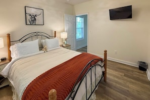 Quarto com cama, mesa de cabeceira, luminária, quadro pendurado na parede e televisão fixada na parede.