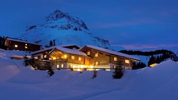 Uma pousada de montanha coberta de neve, com várias janelas e iluminação externa ao entardecer.