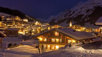 Vila montanhosa coberta de neve à noite, com edifícios iluminados e uma pousada destacada com varanda.