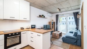 Cozinha compacta com armários brancos, forno embutido e micro-ondas. Há uma pequena área de refeições com mesa e cadeiras, e uma sala de estar com sofá e vista através de uma porta de vidro.
