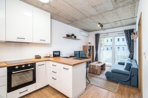 Cozinha compacta com armários brancos, forno embutido e micro-ondas. Há uma pequena área de refeições com mesa e cadeiras, e uma sala de estar com sofá e vista através de uma porta de vidro.