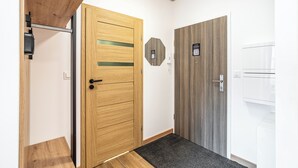 Um corredor com duas portas, uma com acabamento em madeira e outra com acabamento cinza. Há um tapete preto na entrada e uma parede branca com um interruptor de luz.