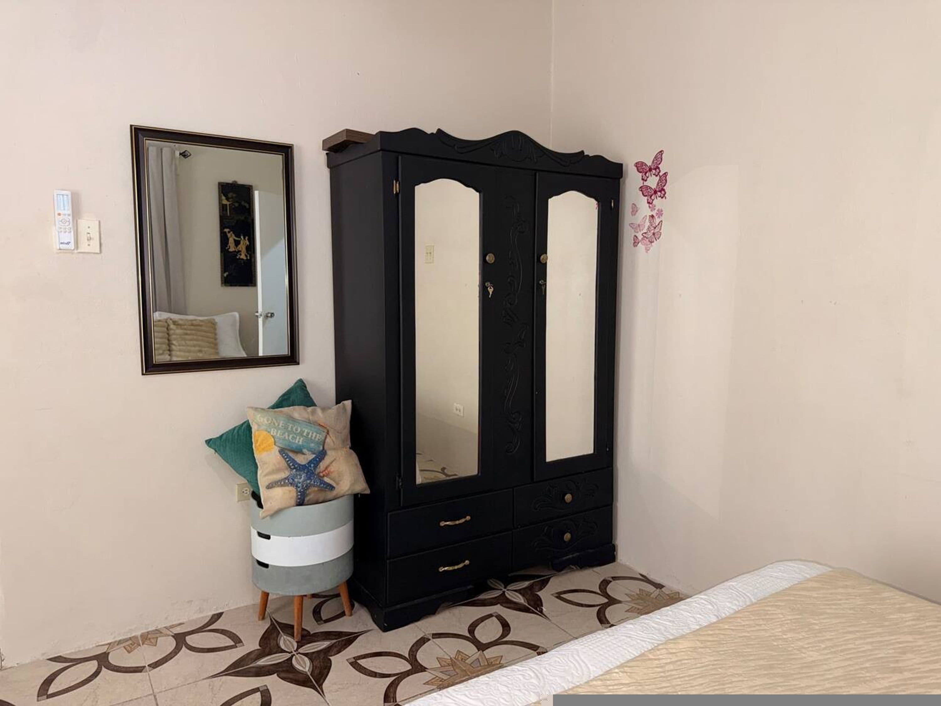 Quarto com cama, guarda-roupa, espelho e um pequeno banquinho com almofada.