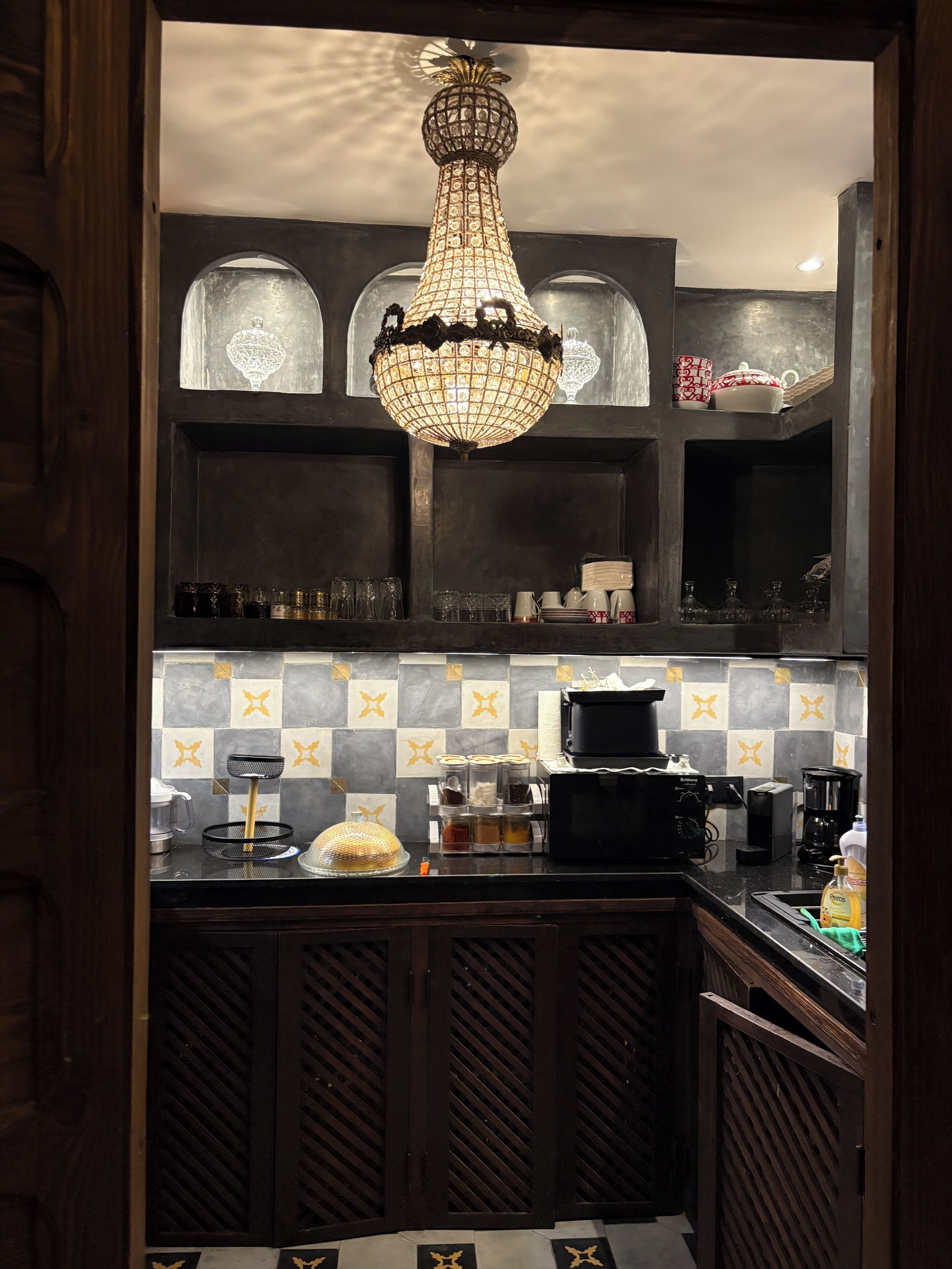 Uma cozinha com um candelabro, prateleiras com copos de vidro, uma cafeteira e um painel de fundo padronizado.